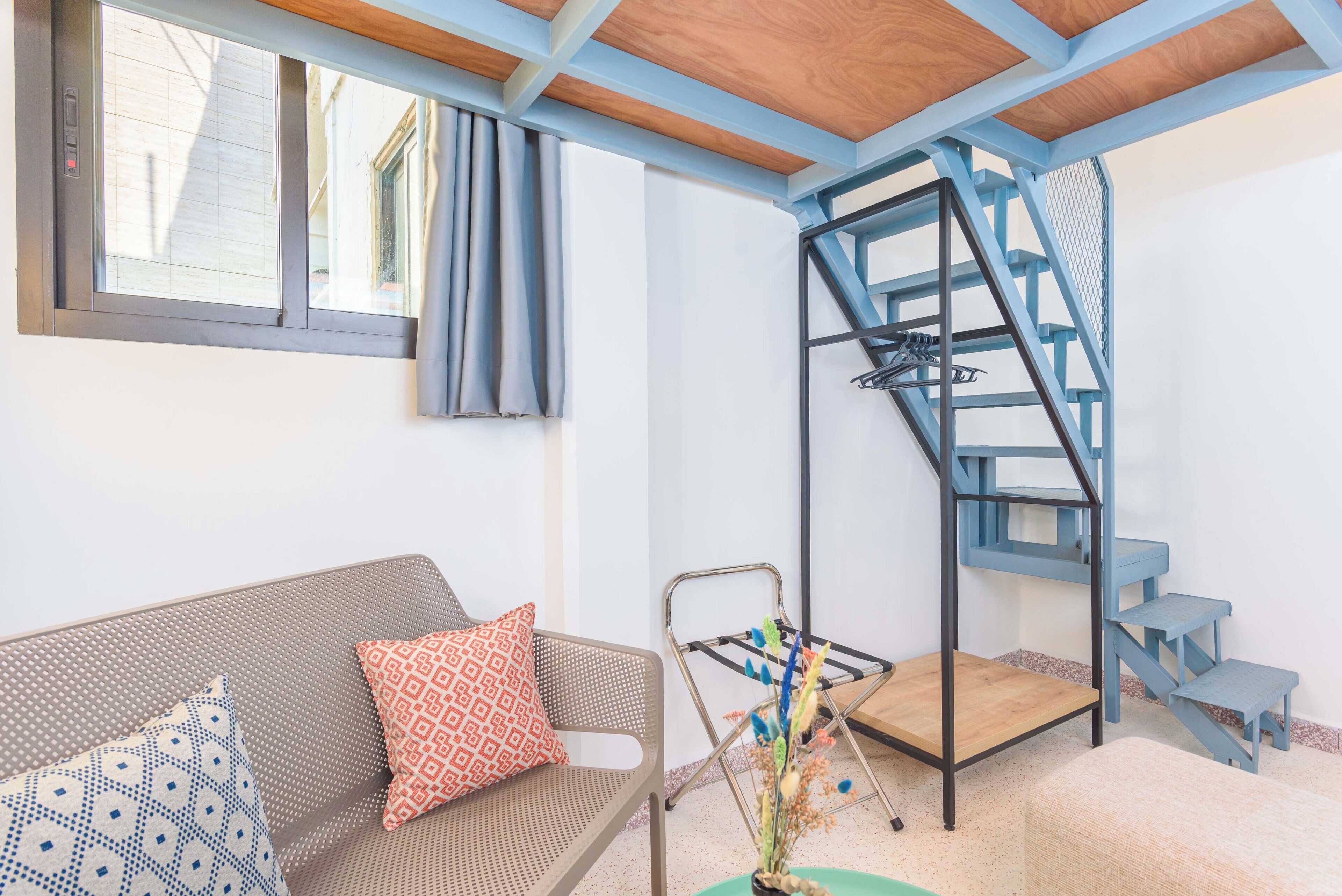 Quarto com sofá, uma mesinha com um vaso, uma cadeira de metal e uma escada azul.