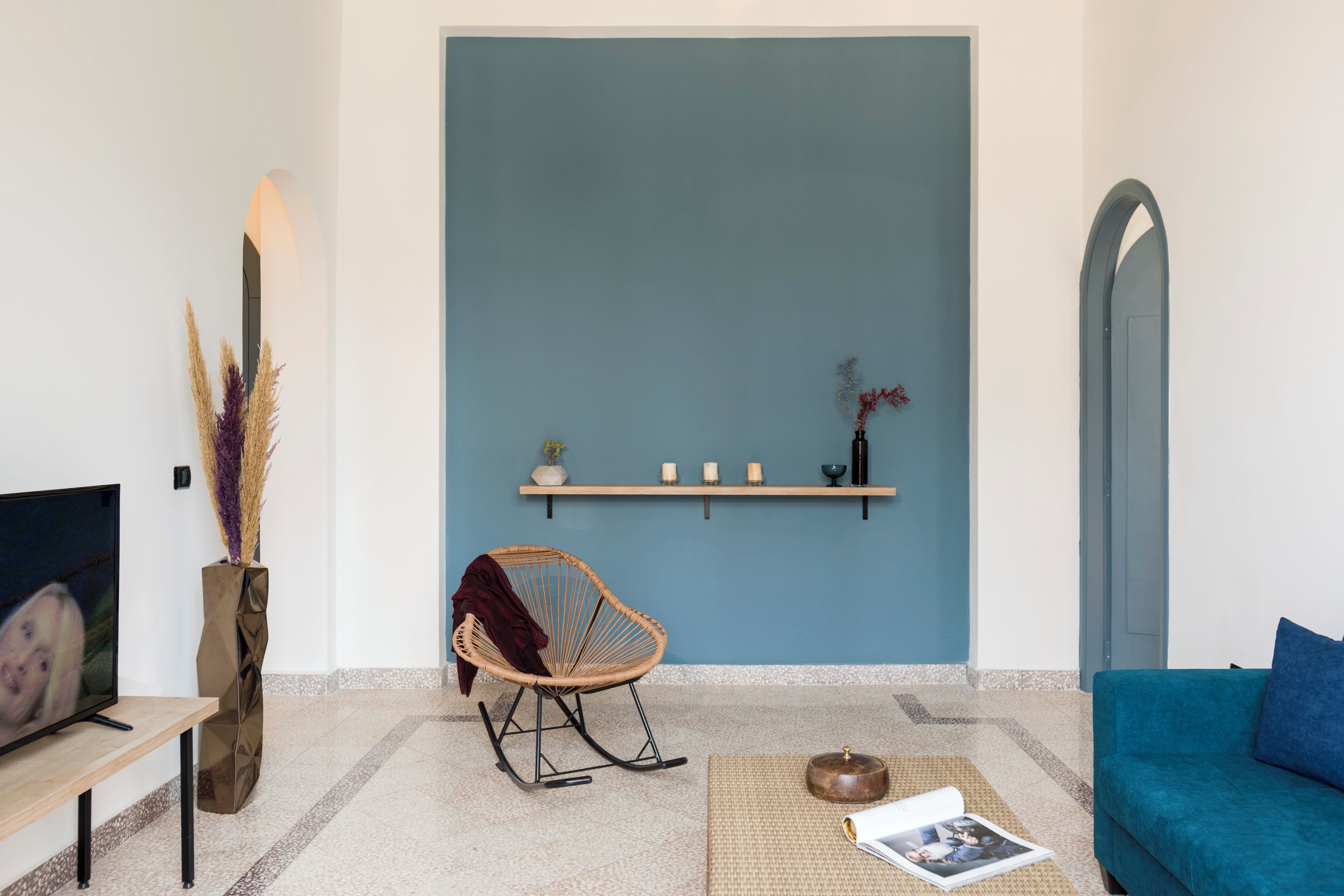 Uma sala de estar com televisão, uma poltrona de balanço, um sofá azul, uma estante de livros e um vaso decorativo.