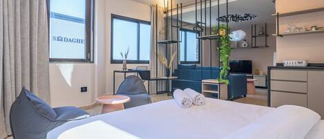 Quarto de hotel moderno com uma cama grande, uma escrivaninha, uma cadeira e um sofá. Há uma janela com cortinas, uma planta e uma televisão.