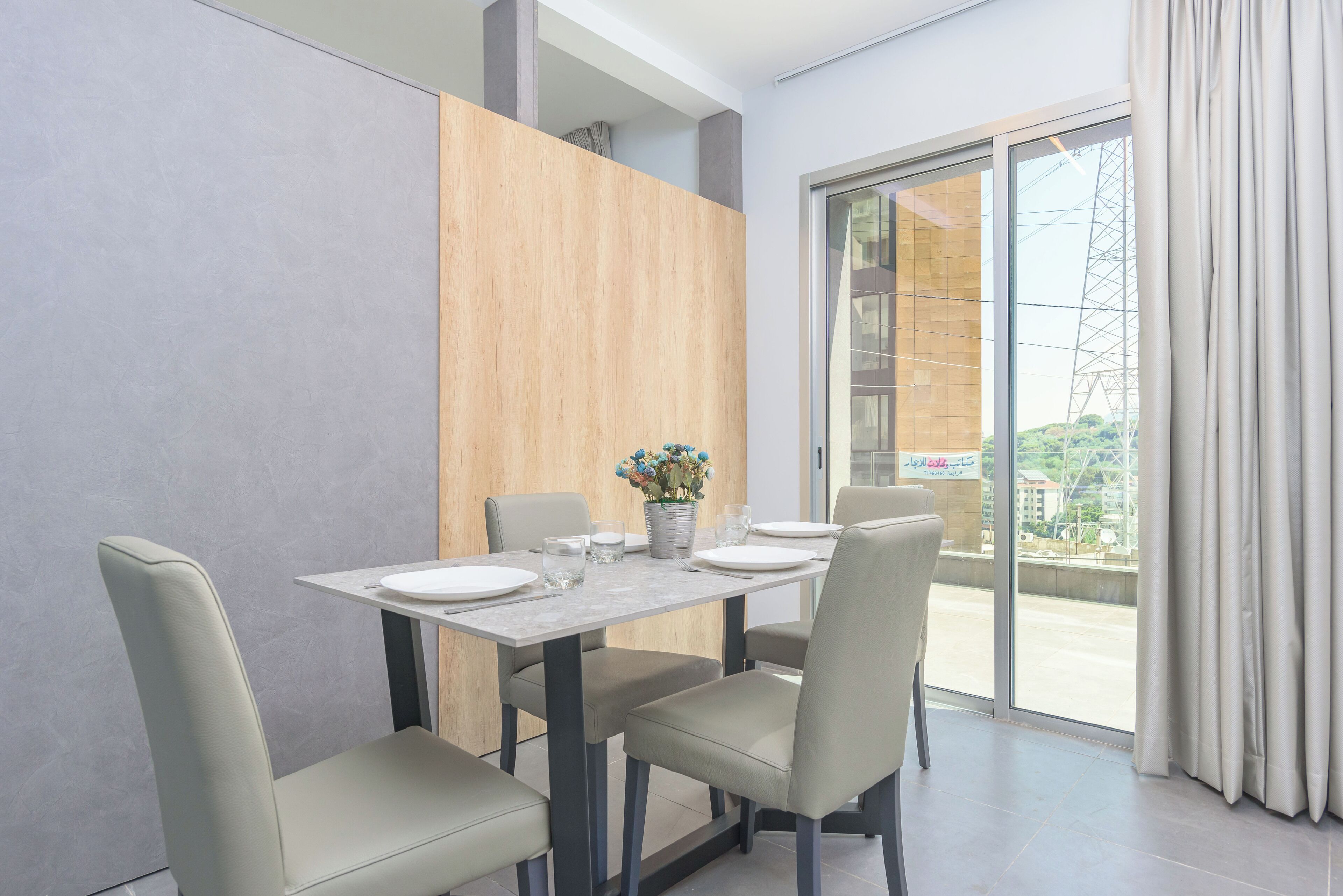 Uma sala de jantar com uma mesa posta para quatro pessoas, cadeiras, um vaso com flores e vista para um edifício através de uma porta de vidro.