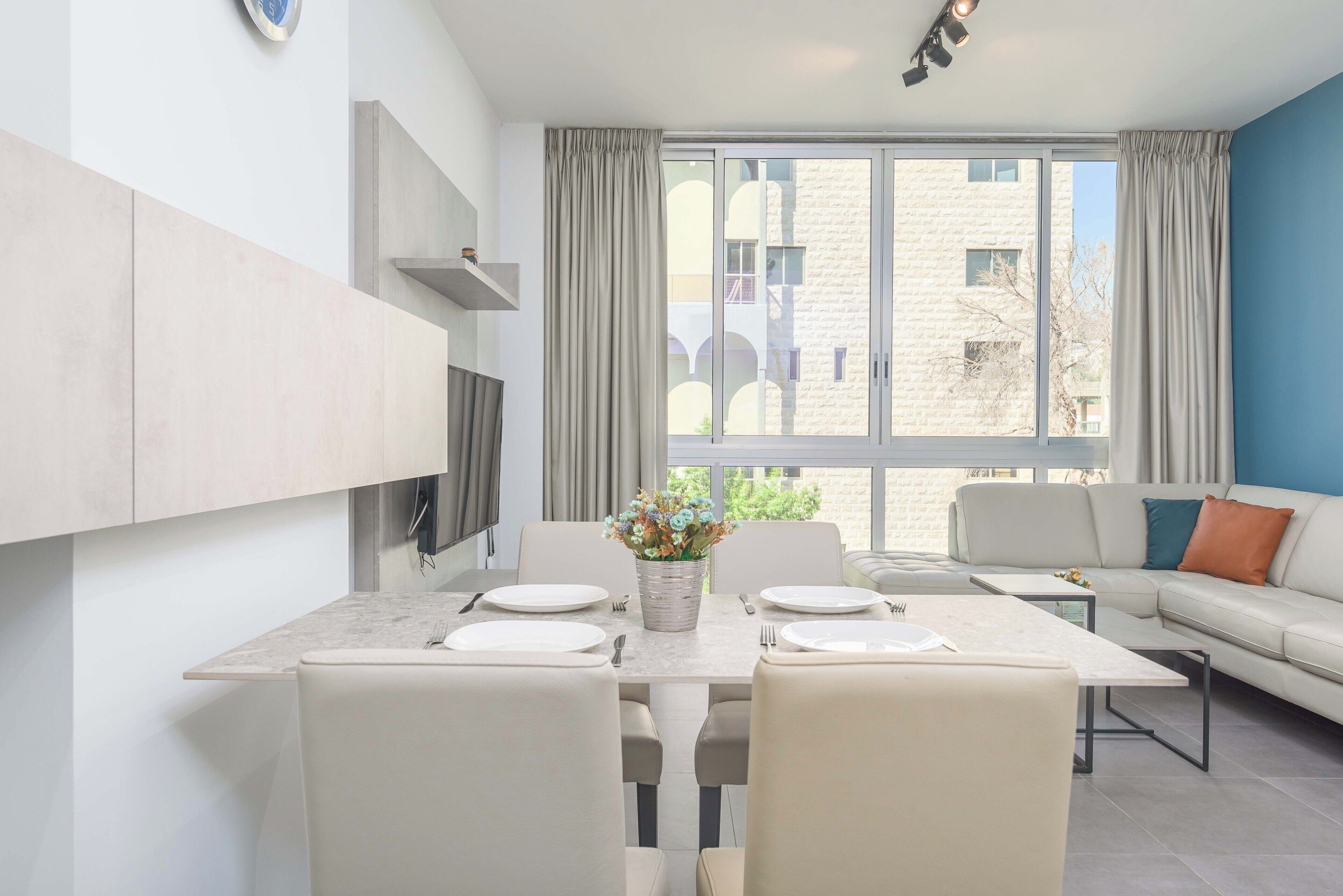 Uma sala de jantar moderna com uma mesa posta para quatro pessoas, um sofá e uma grande janela com cortinas.