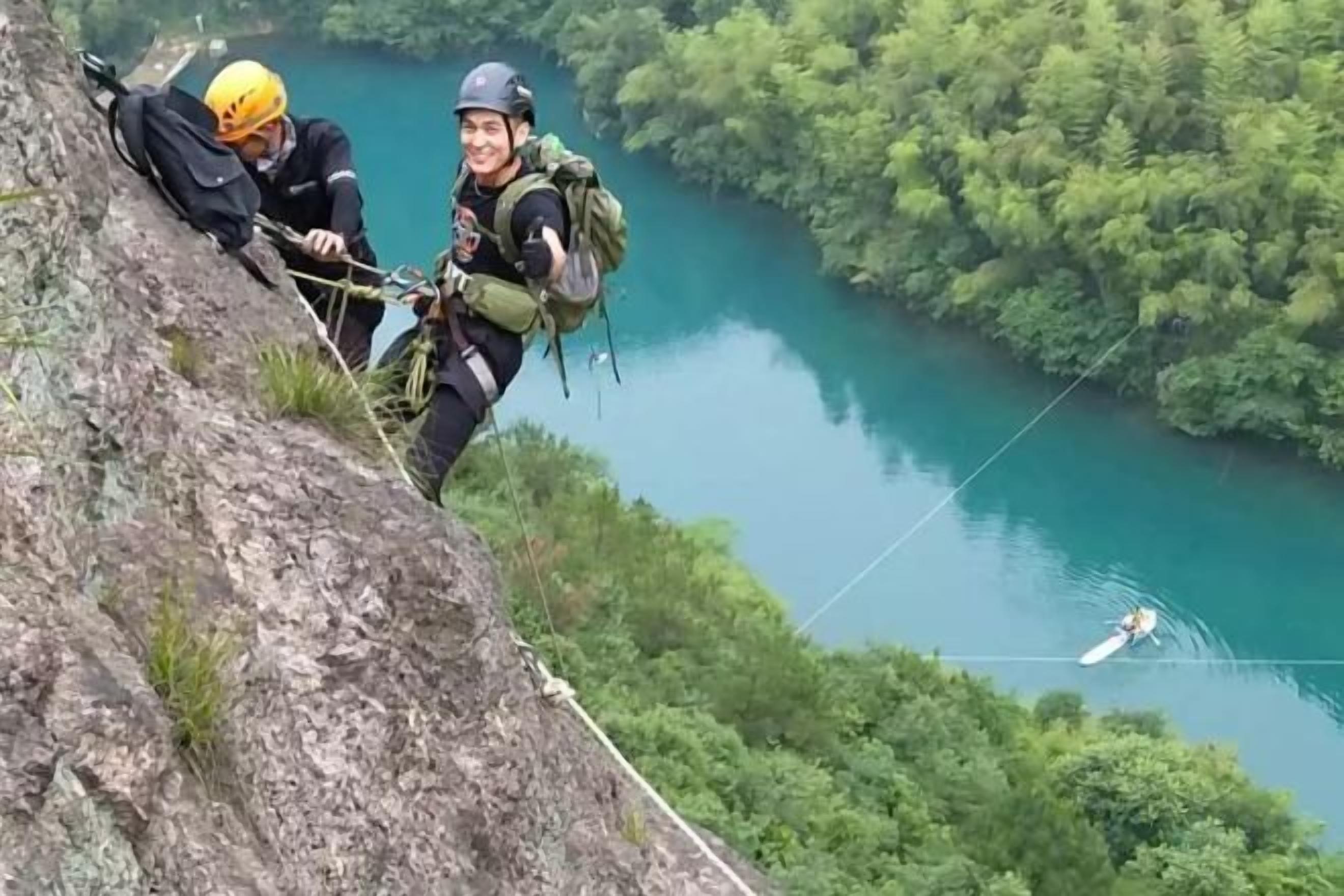 Duas pessoas fazem rapel em um penhasco, com um rio e uma floresta ao fundo.