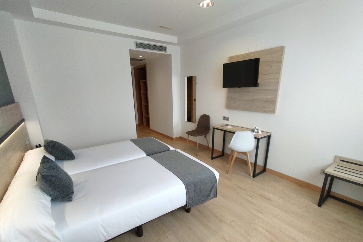 Quarto de hotel moderno com uma cama grande, uma escrivaninha, uma cadeira e uma televisão na parede.