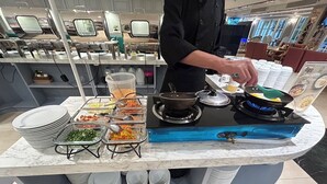 Disposição de buffet com diversos alimentos, um fogão e uma pessoa cozinhando.