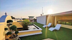 Jardim no terraço com grama sintética, um pequeno lago, plantas em vasos e cadeiras de lounge brancas.