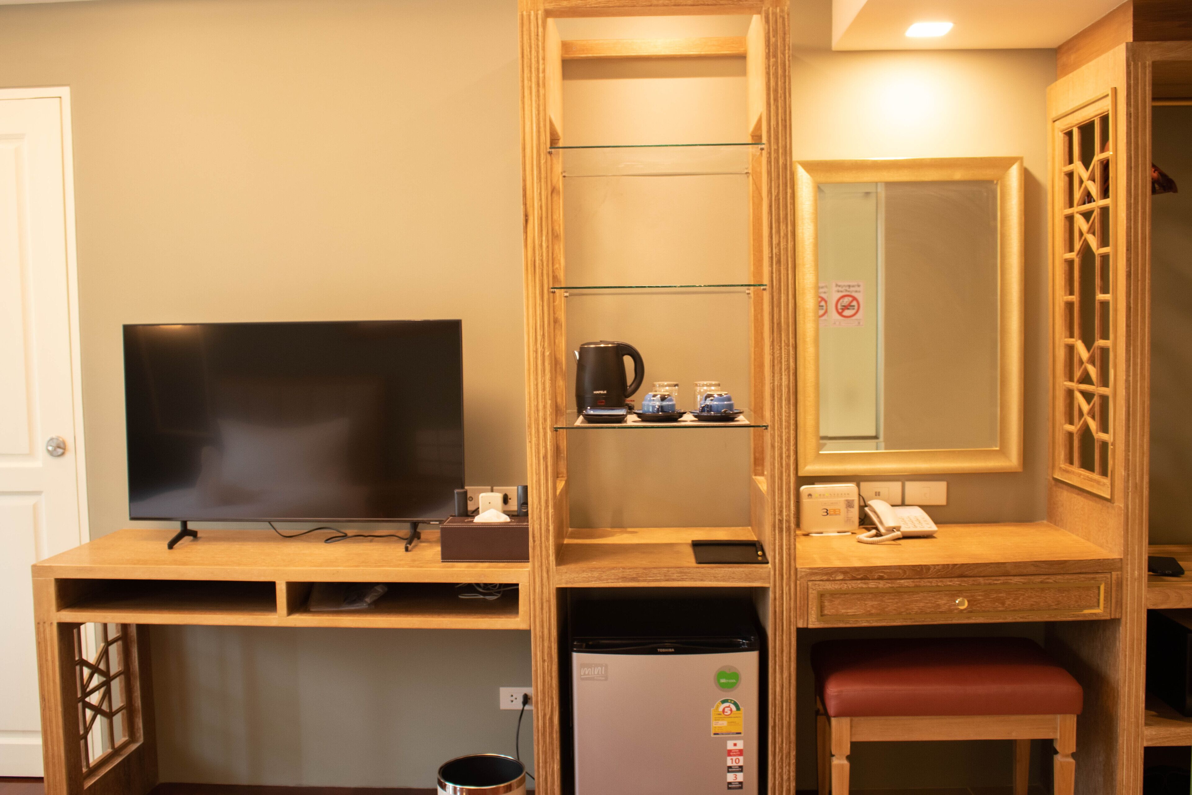 Quarto compacto com escrivaninha de madeira, televisão, máquina de café, mini-geladeira e um pequeno banco.