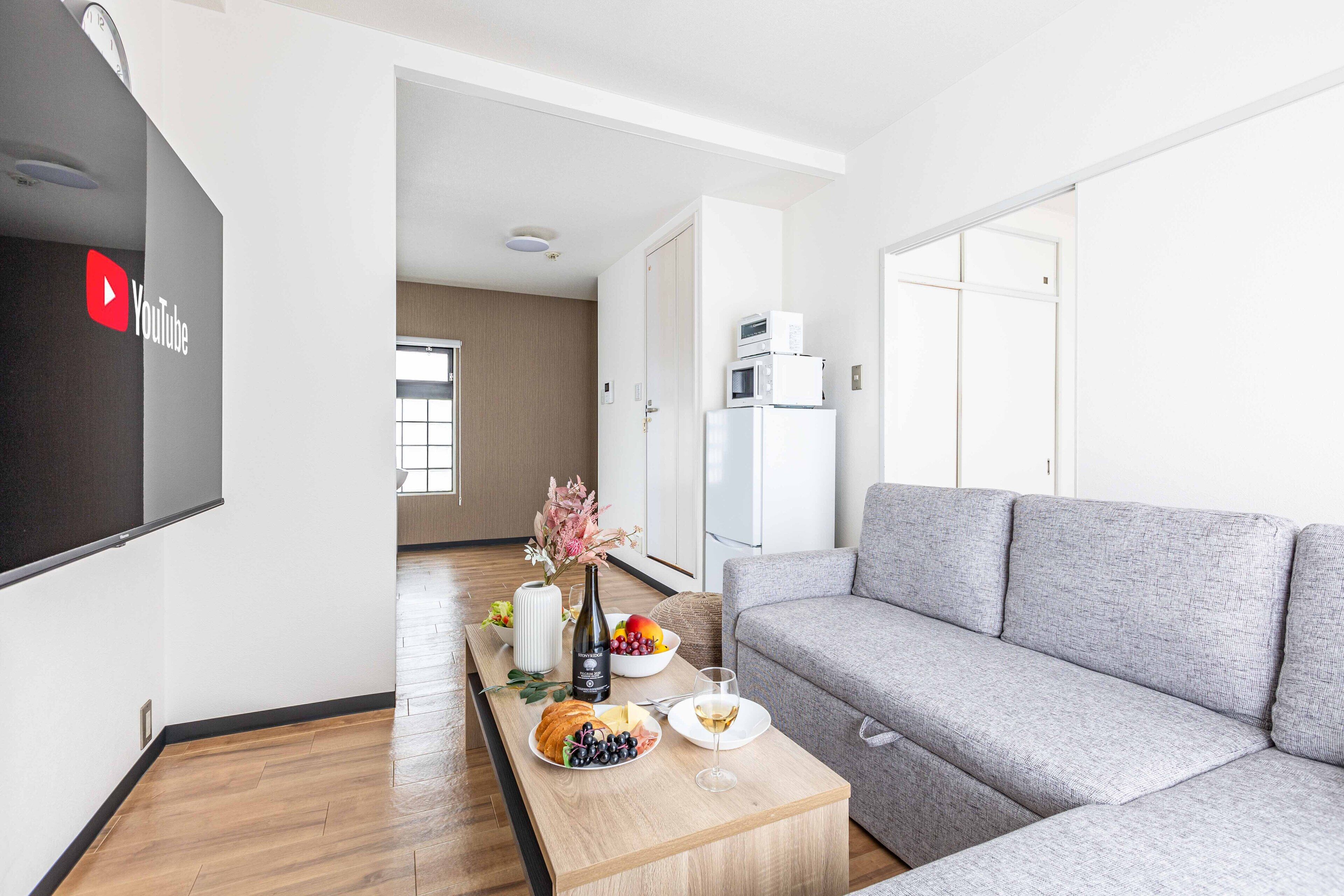 Uma sala de estar moderna com um sofá cinza, uma mesa de centro de madeira com comida e bebidas, uma televisão de tela plana e uma área de cozinha com micro-ondas.