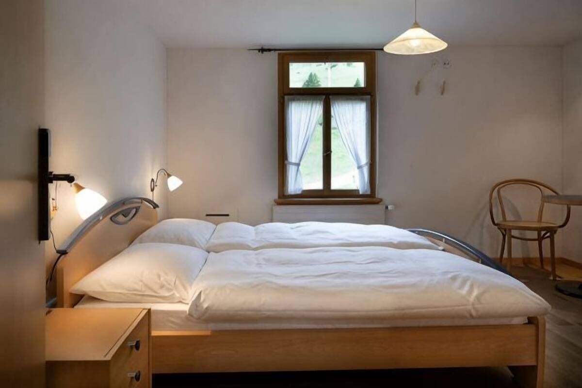 Quarto com cama, mesa de cabeceira, abajur, janela e cadeira.