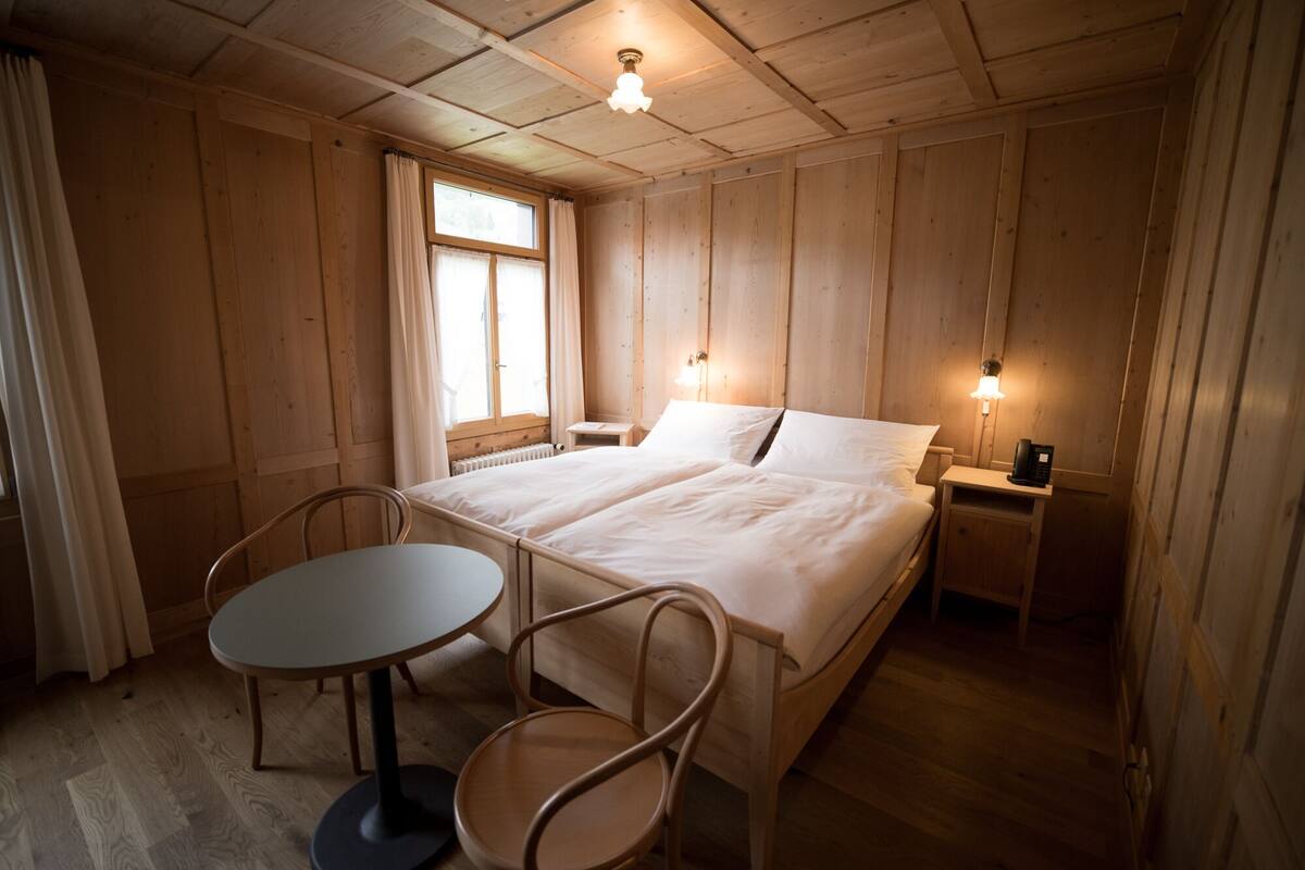 Um quarto de madeira com uma cama, duas cadeiras, uma mesa, uma janela com cortinas, um abajur e um telefone.