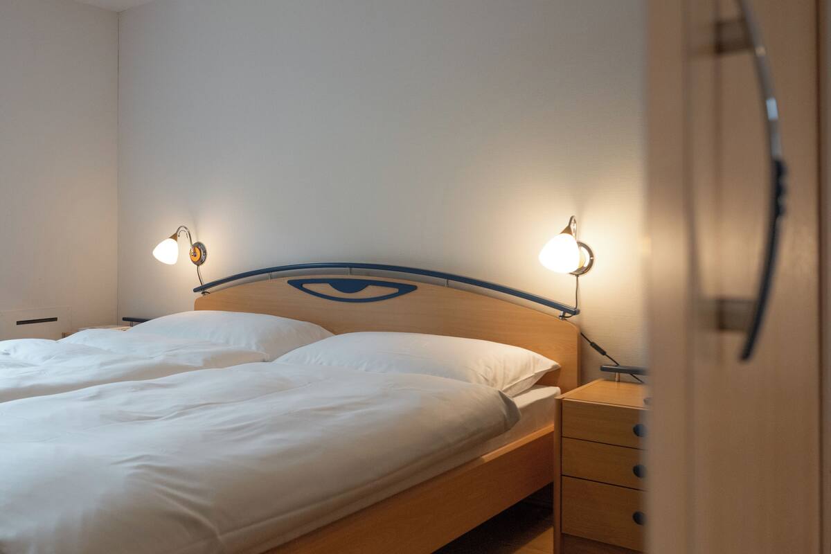 Uma cama com cabeceira de madeira, duas mesas de cabeceira e um abajur fixado na parede.