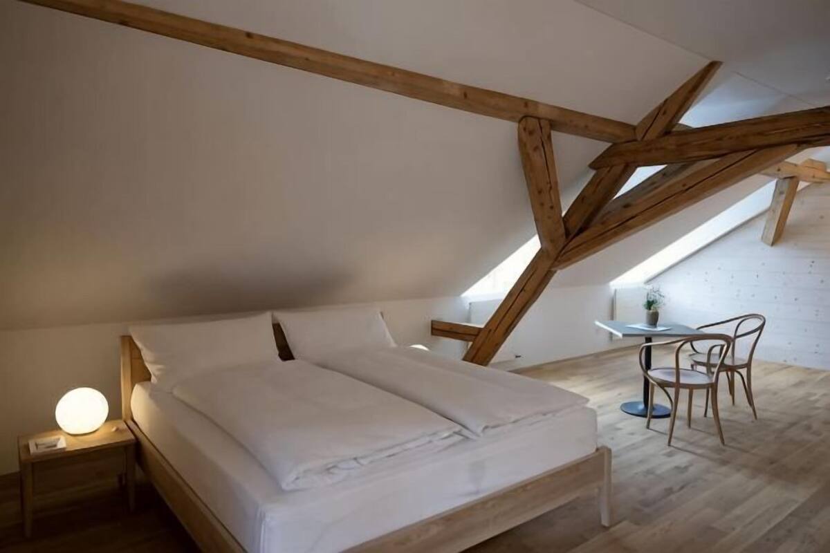 Quarto com teto de madeira, uma cama, mesa de cabeceira com abajur e uma pequena mesa com cadeiras.