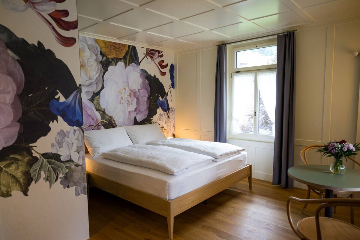 Quarto com cama de madeira, um mural floral na parede, uma janela com cortinas e uma pequena mesa com um vaso de flores.