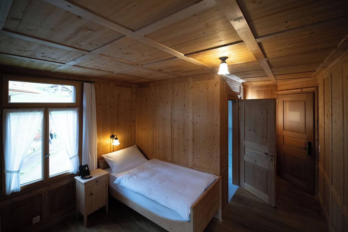 Um quarto de madeira com uma cama, uma mesa de cabeceira, uma janela com cortinas e uma porta.