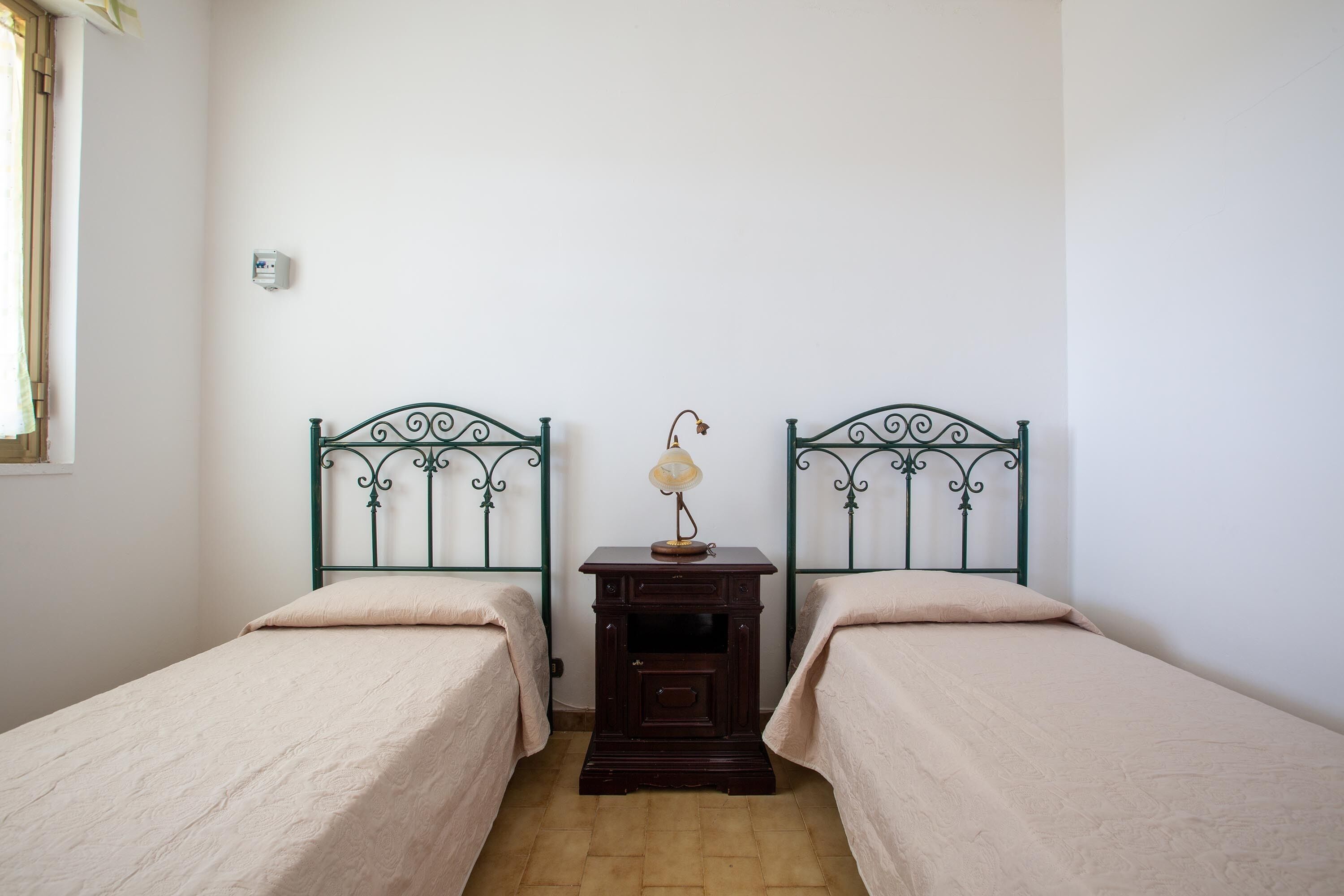 Quarto com duas camas de solteiro, mesa de cabeceira com abajur e tomada na parede.