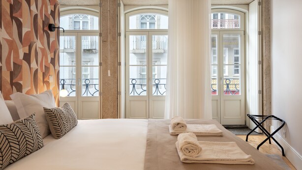 Quarto de hotel com uma cama grande, duas janelas e vista para um edifício com varandas.