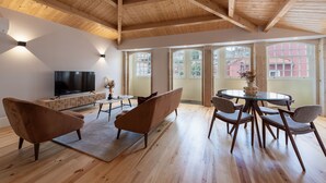 Ampla sala de estar com teto de madeira alto, TV de tela plana, sofá, mesa de jantar com cadeiras e grandes janelas com vista para edifícios.