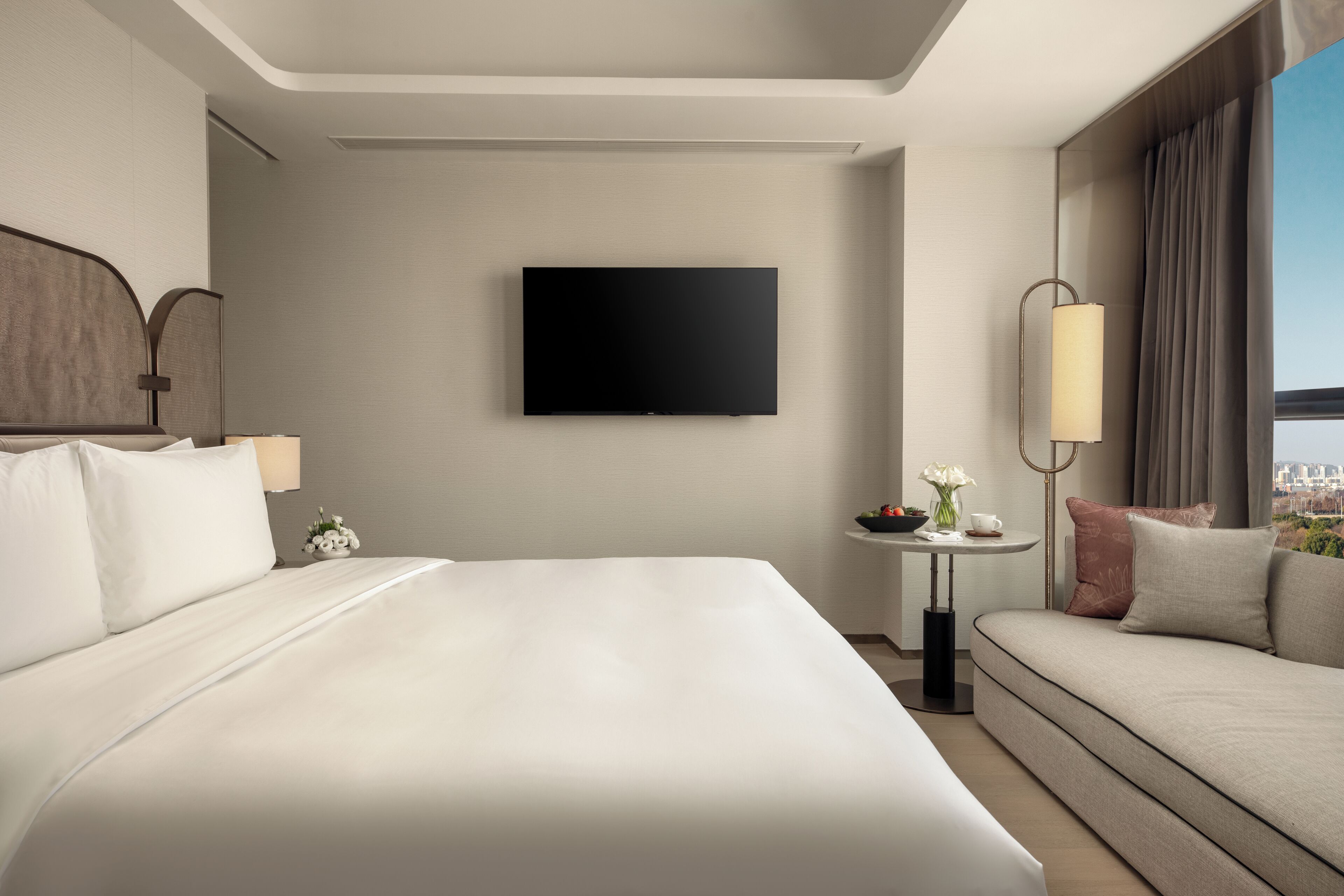 Quarto de hotel moderno com uma cama grande, um sofá, uma TV de tela plana na parede, uma mesa pequena com um vaso e uma xícara, e vista da cidade pela janela.