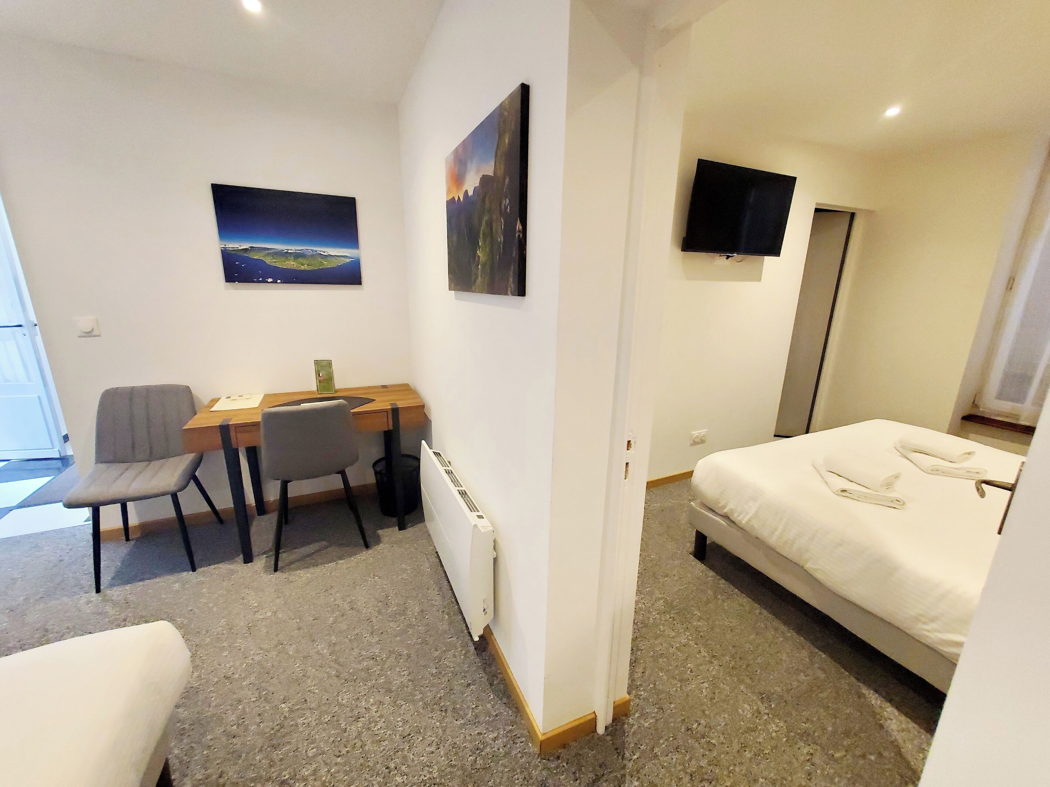 Quarto de hotel com uma cama, uma escrivaninha com cadeira, uma televisão na parede e dois quadros.