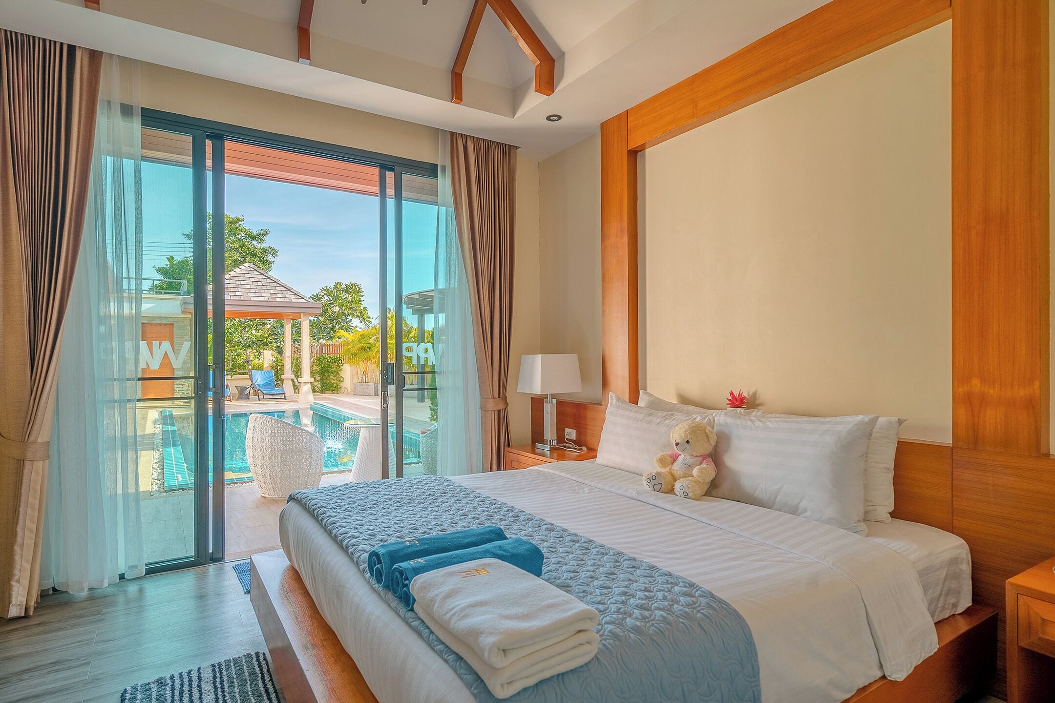 Quarto de hotel com cama, mesa de cabeceira, luminária, um ursinho de pelúcia e vista para piscina e área externa.