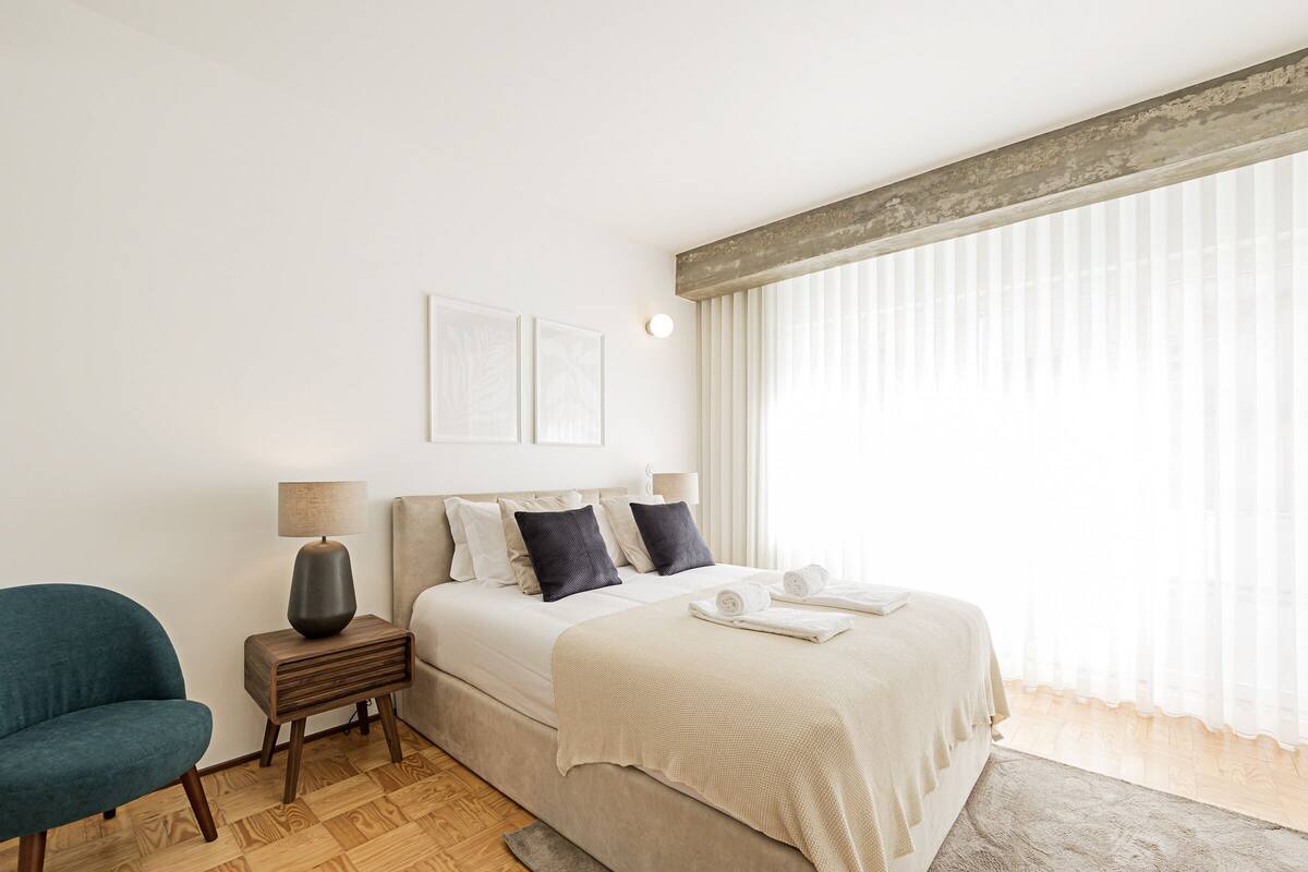Quarto de hotel com cama, cadeira, mesa de cabeceira e janela com persianas.