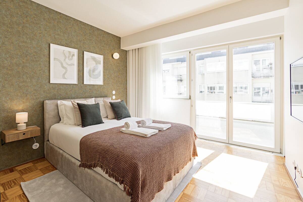 Quarto moderno com uma cama grande, mesa de cabeceira com abajur e vista da cidade através de grandes janelas.