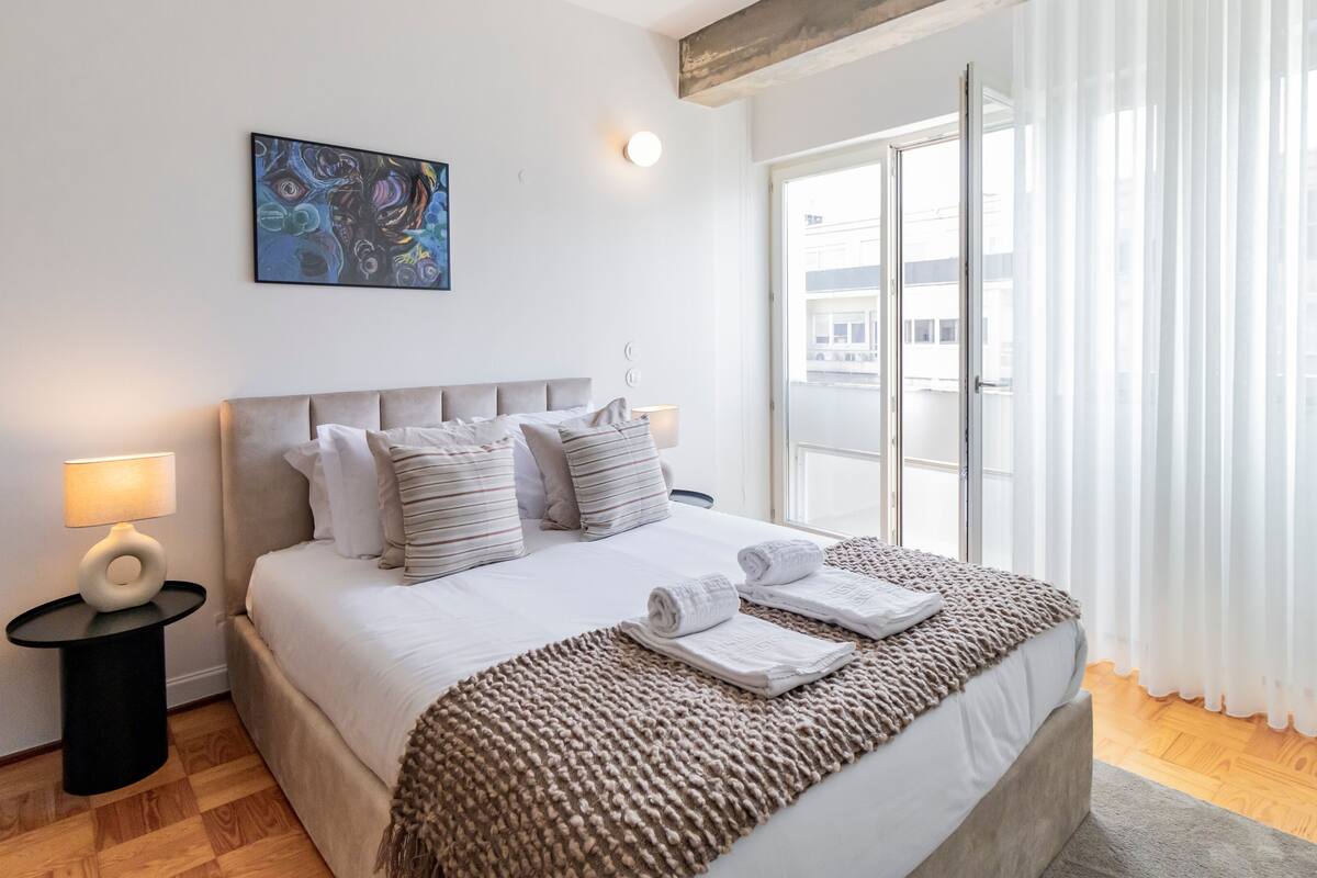 Quarto moderno com uma cama grande, uma mesinha de cabeceira com abajur, um quadro na parede e vista da cidade pela janela.