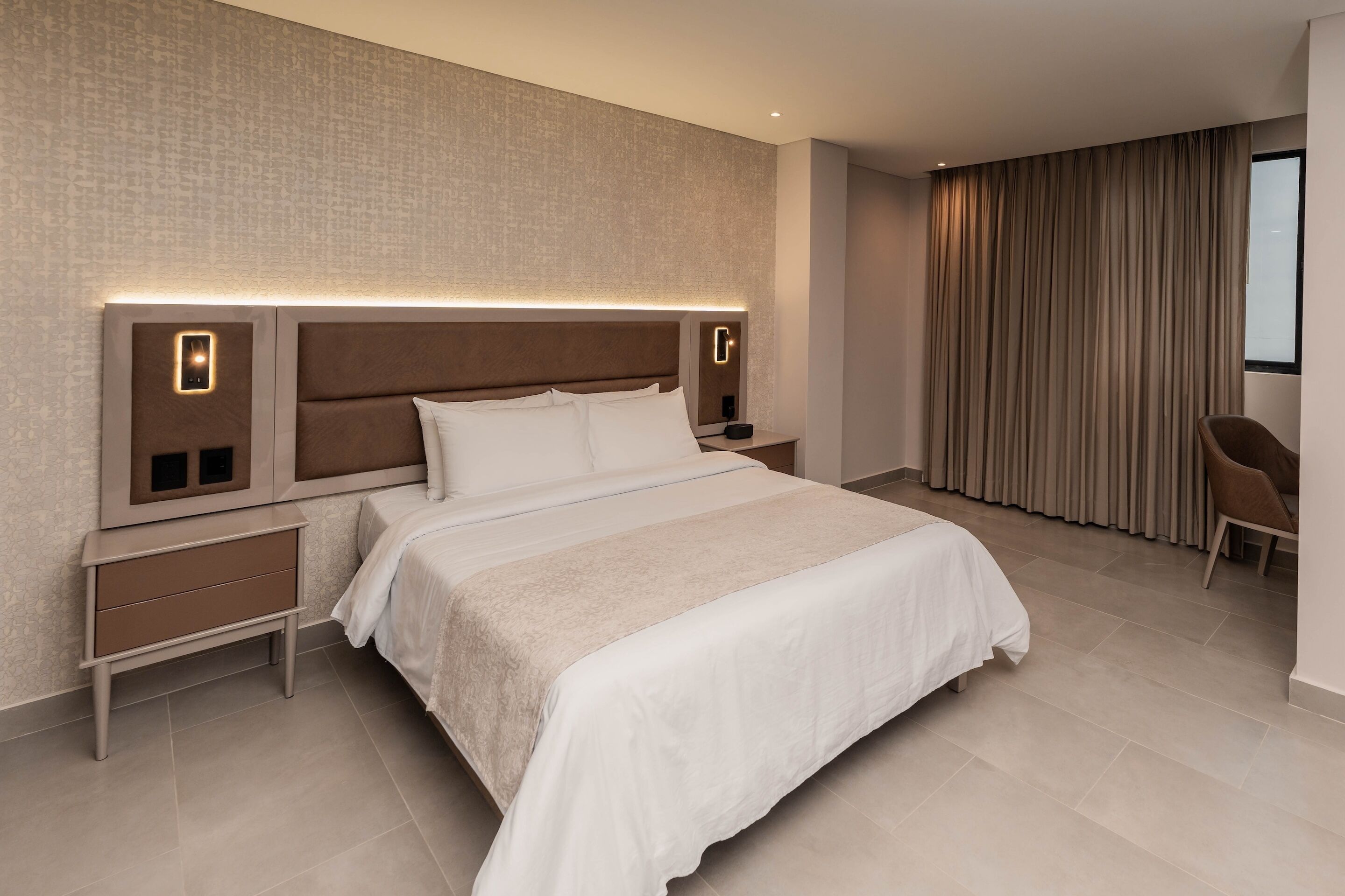 Quarto de hotel moderno com uma cama grande, mesas de cabeceira, uma escrivaninha e uma cadeira.