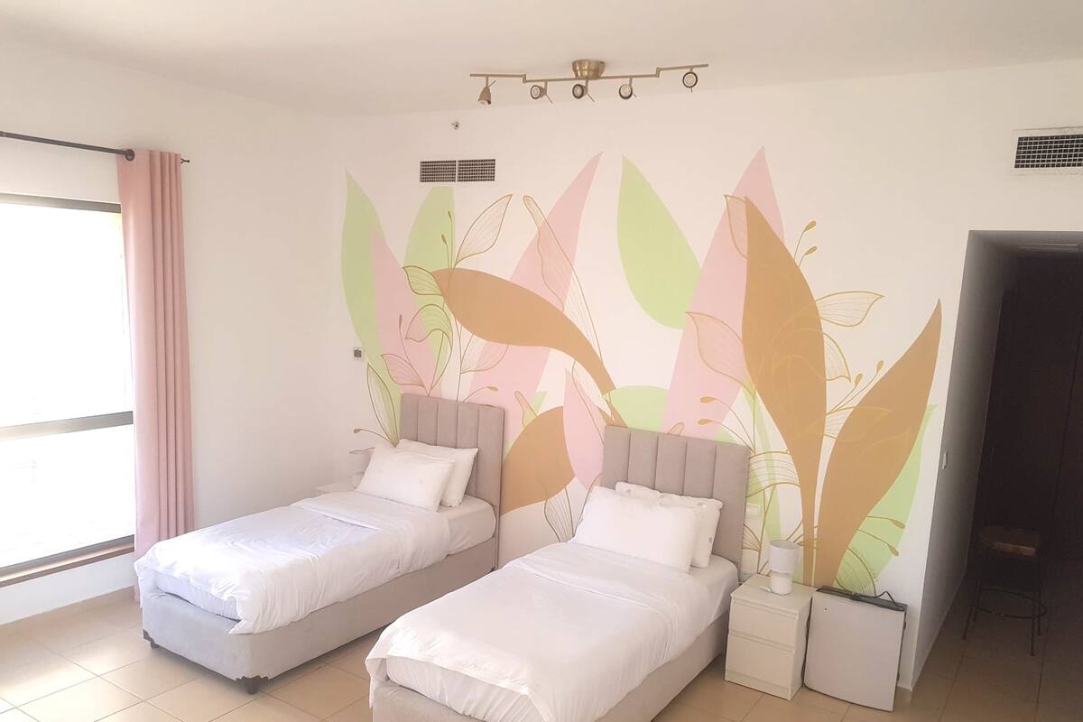 Quarto com duas camas, janela com cortinas, parede com padrão de folhagens e luminária no teto.