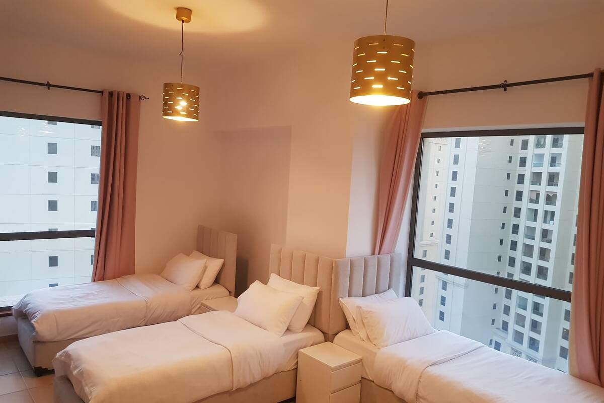 Quarto de hotel moderno com duas camas, uma mesinha de cabeceira e grandes janelas com vista para a cidade.