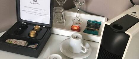 Quarto de hotel com cafeteira, bandeja com máquina Nespresso, xícara, pires, duas taças de vinho e um objeto preto que parece um apoio para os pés.