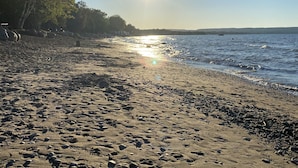 Uma praia com pegadas visíveis e pedras, árvores à esquerda, e o sol refletindo na água.