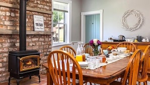 Uma sala de jantar com parede de tijolos, uma lareira a lenha preta, cadeiras de madeira, uma mesa posta para refeição e um espaço de cozinha com uma porta azul e uma coroa na parede.