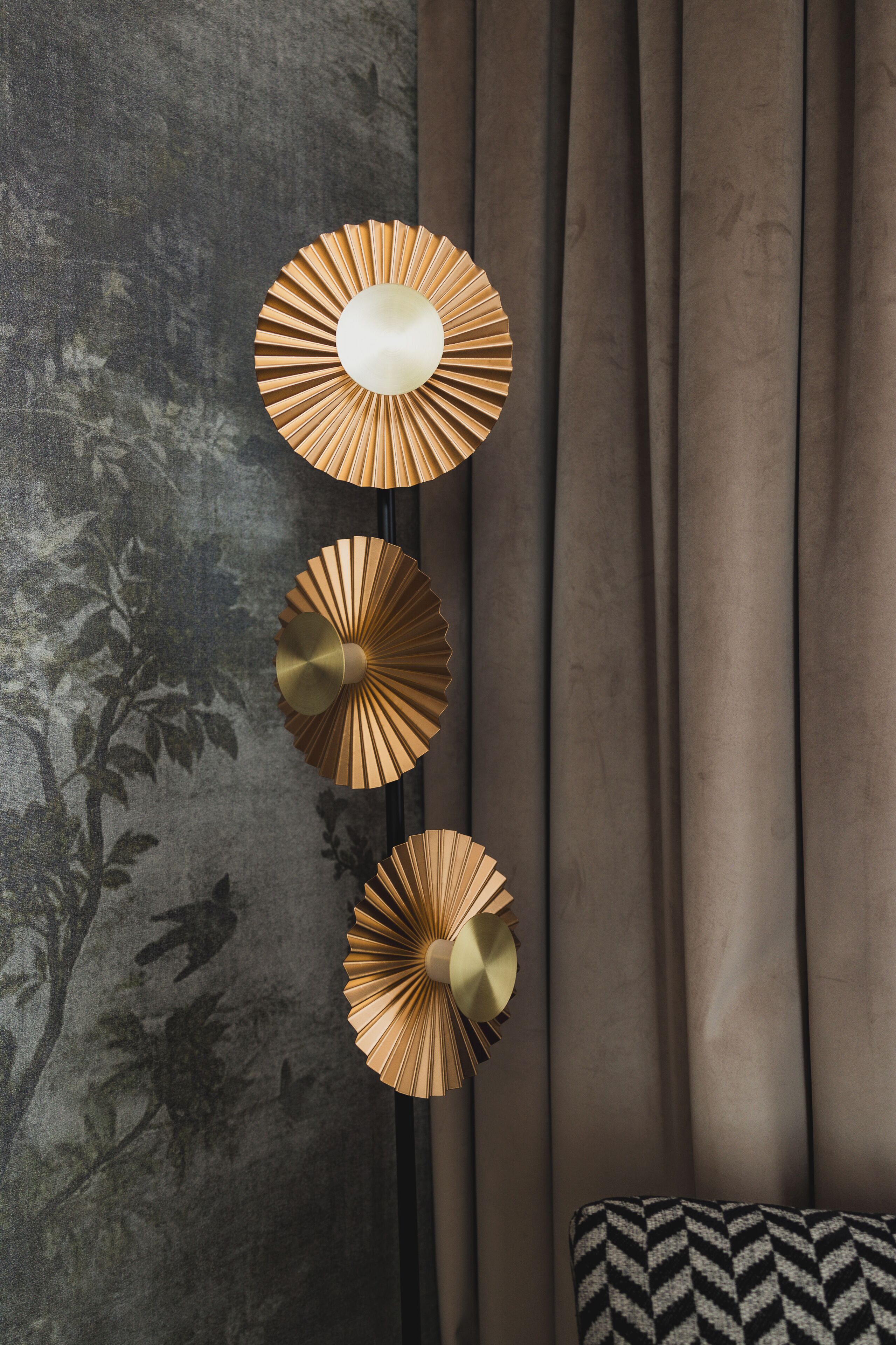 Luminária circular dourada, fixada na parede, com vários níveis e um elemento refletor central, em frente a uma parede texturizada com padrão floral e cortinas escuras.