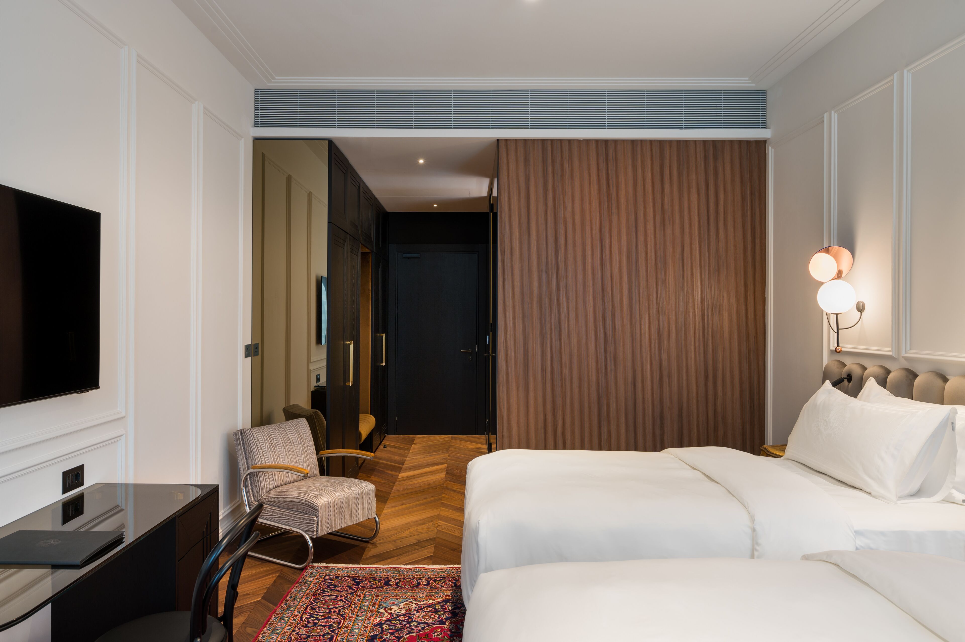 Quarto de hotel com cama, escrivaninha, cadeira, televisão e porta que dá para um corredor.
