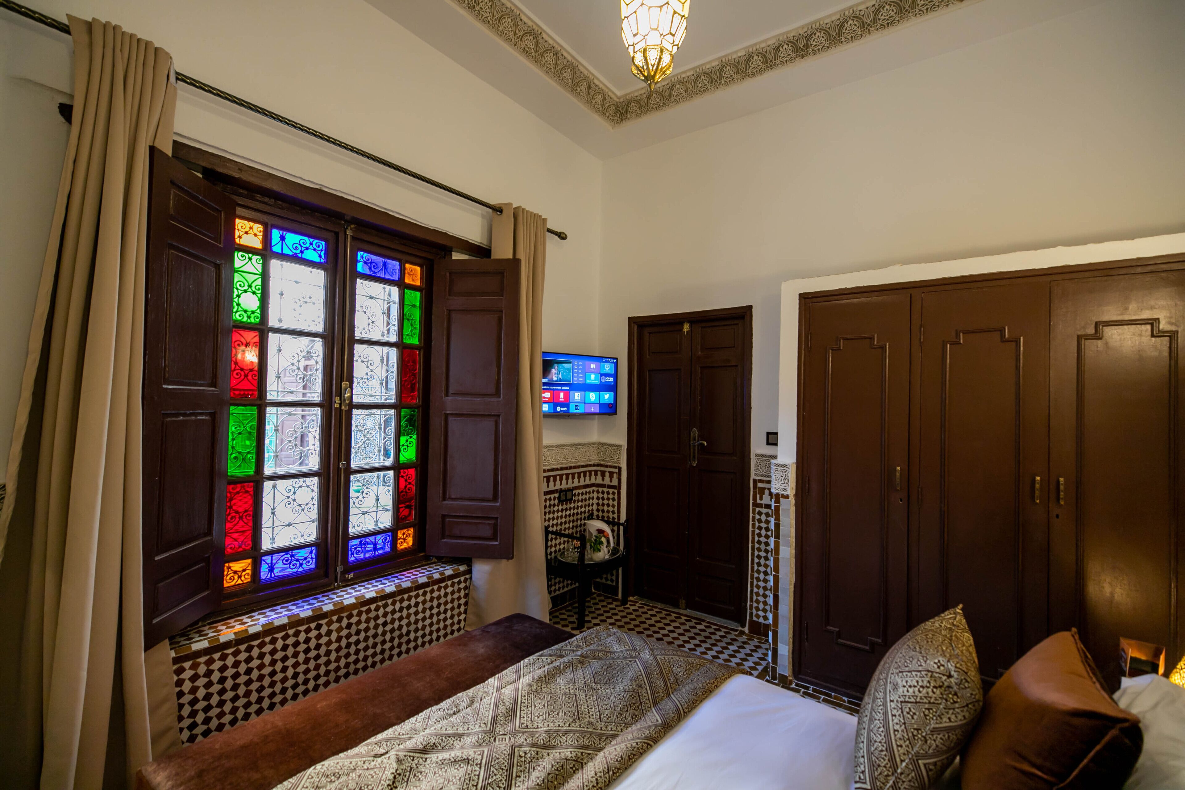 Quarto com cama, televisão na parede, guarda-roupa e janela com vitrais.