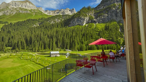 Um terraço com cadeiras e mesas vermelhas, com vista para um campo de golfe e montanhas.