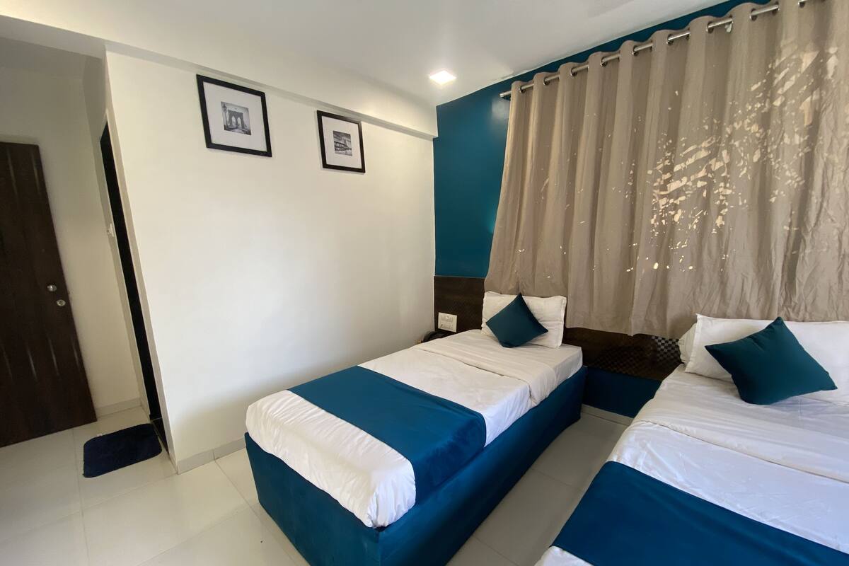 Quarto de hotel com duas camas de solteiro, uma janela grande com cortinas e uma porta à esquerda.