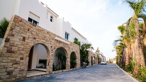 Rua pavimentada com edifícios brancos, arcos de pedra e palmeiras.