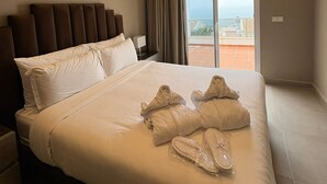 Quarto de hotel com uma cama grande, vista da cidade através de uma porta de correr e uma pintura de pôr do sol na parede.