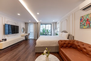 Quarto de hotel moderno com uma cama grande, um sofá de couro marrom, uma TV, uma mesa de trabalho e vista para o verde pela janela.