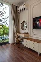 Um quarto com escrivaninha de madeira, uma cadeira, penteadeira, televisão e janela com cortinas.