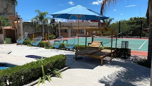 Uma área de piscina com espreguiçadeiras, quadra de tênis e um edifício ao fundo.