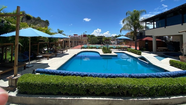 Piscina com espreguiçadeiras e guarda-sóis, rodeada por vegetação e edifícios.