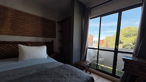 Quarto com cama grande, cabeceira de madeira, vista da cidade através de uma janela ampla e uma mesinha perto da janela.