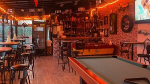 Mesa de bilhar, área de bar e disposição de assentos em um espaço com paredes de tijolo e piso de madeira.