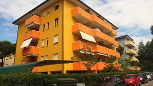 Um edifício amarelo de vários andares com varandas laranjas, rodeado por carros estacionados e vegetação.