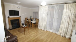 Uma sala de estar com lareira, uma mesa de jantar com cadeiras e uma janela grande com cortinas.