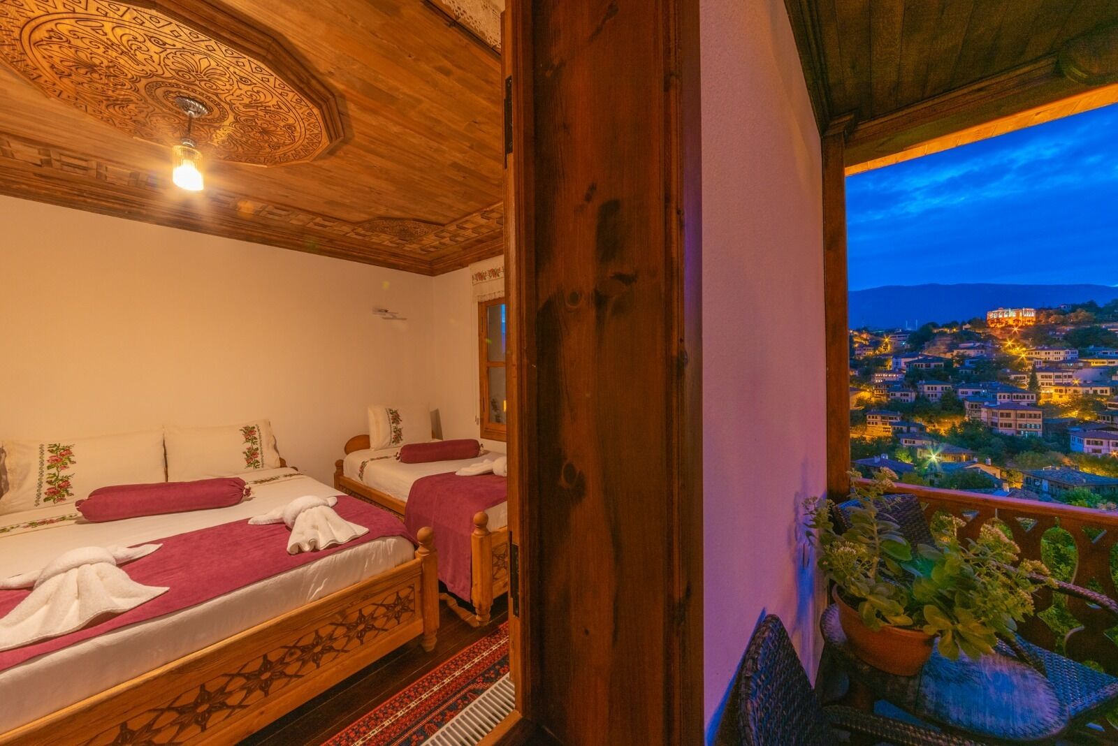 Quarto com duas camas de madeira, teto de madeira ricamente decorado e varanda com vista para uma cidade ao entardecer.