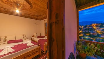 Quarto com duas camas de madeira, teto de madeira ricamente decorado e varanda com vista para uma cidade ao entardecer.