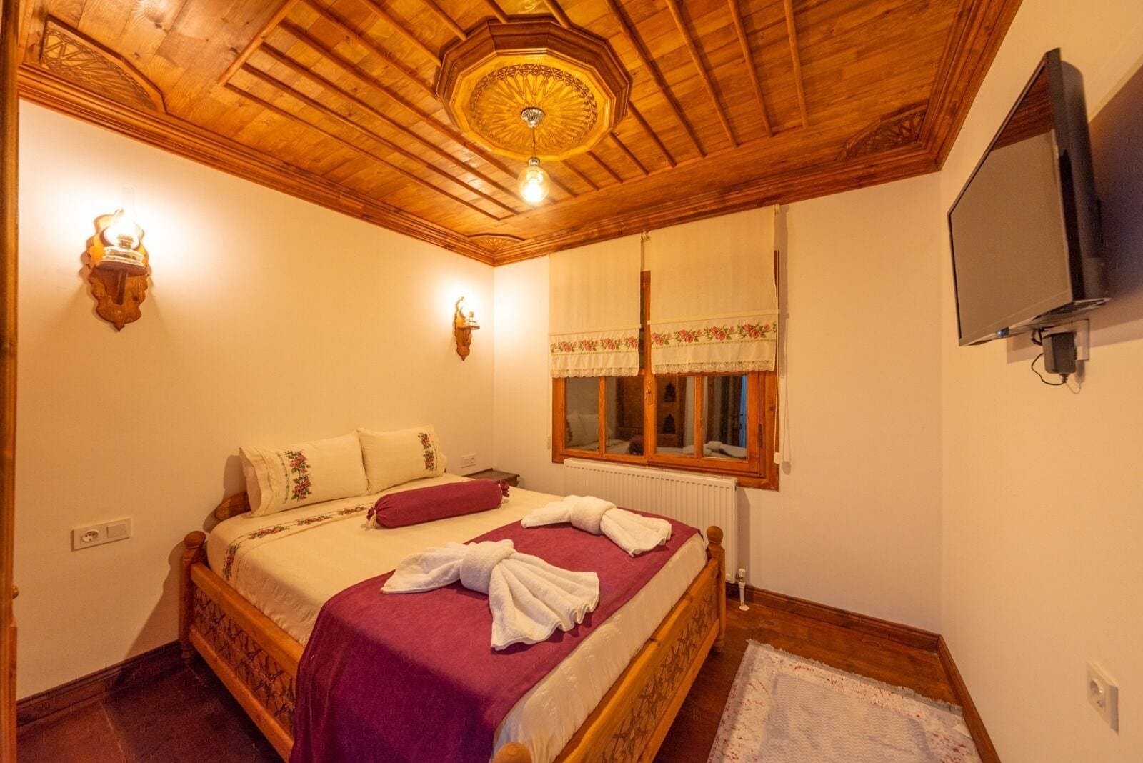 Um quarto aconchegante com teto de madeira, uma cama com roupa de cama florida, uma janela com cortinas e uma TV na parede.